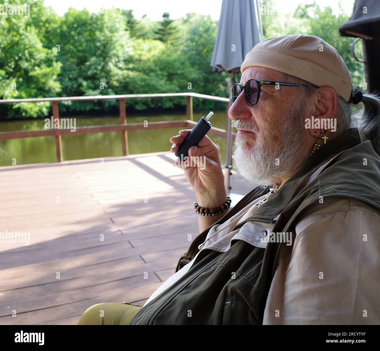 A stout man sits relaxed on a wooden deck terrace and smokes a ...