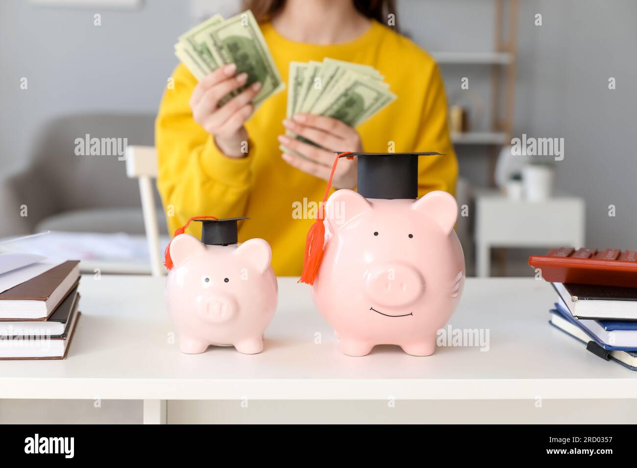 Female student counting money at table with piggy banks in graduation ...