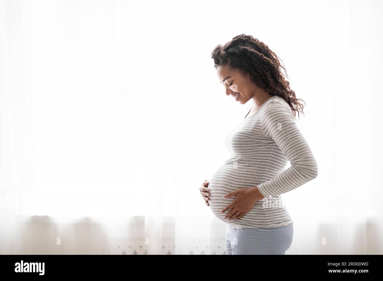 Beautiful pregnant black woman hugging tummy while standing near window ...