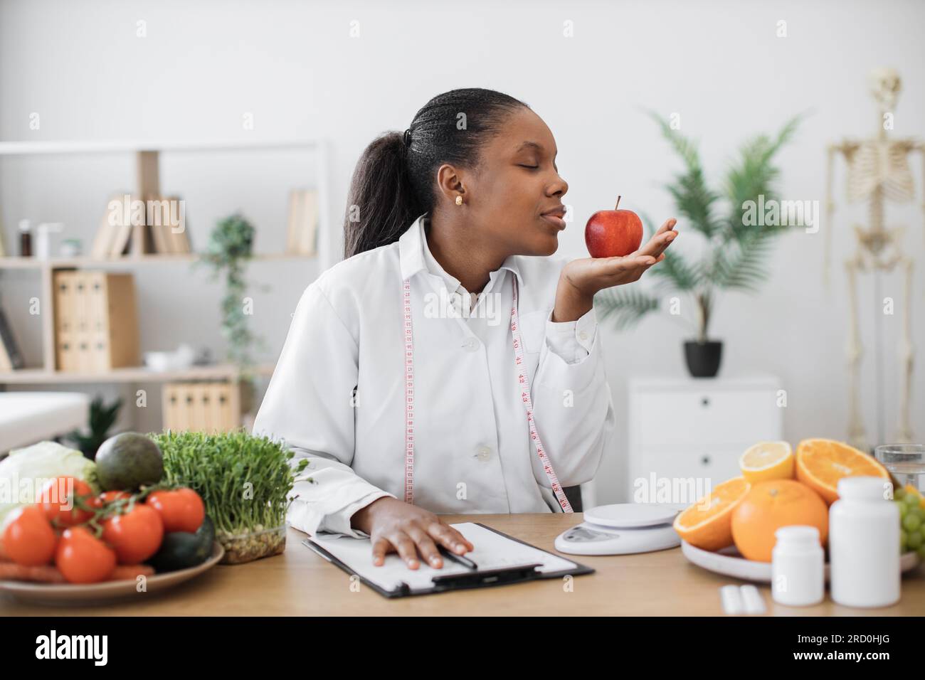 Charming adult lady with measuring tape smelling red apple on palm while noting down nutritional ...