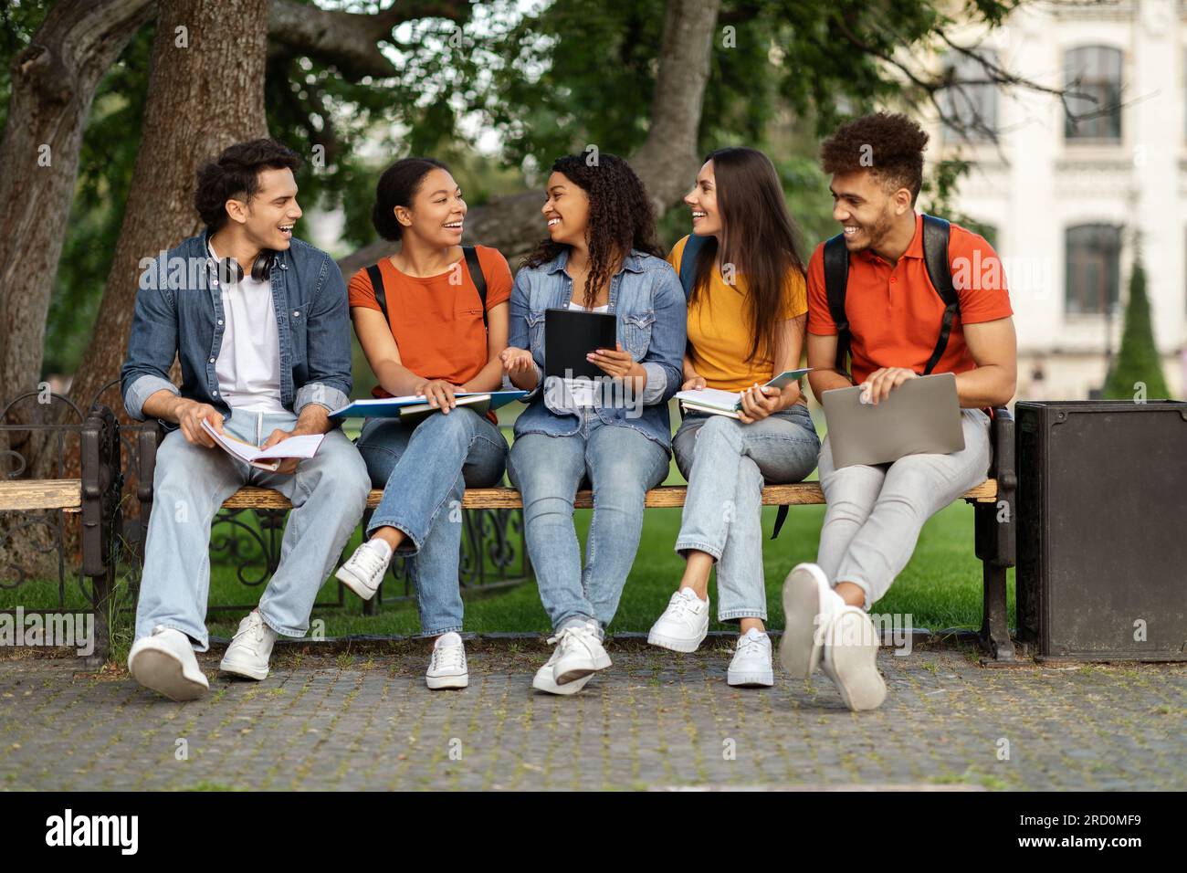 Diverse students friends sitting on bench outdoors in university campus ...