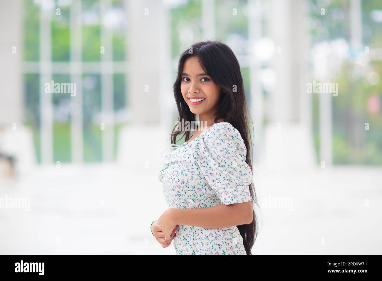Young woman indoors. Girl with long hair wearing a white dress standing at big window in a ...