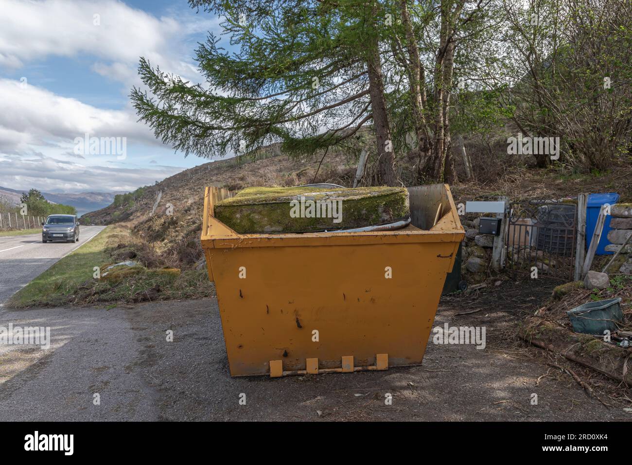 A bright yellow coloured skip with a mattresson the top which is ...