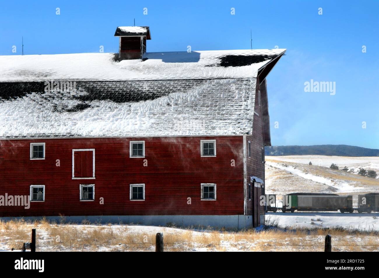 Coal train rumbles by old, wooden red barn. Snow blows from barn roof ...