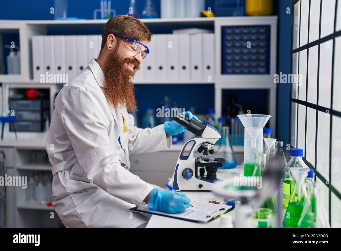 Young redhead man scientist using microscope writing report working at ...