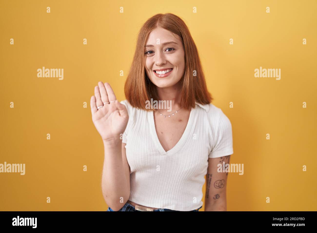 Young redhead woman standing over yellow background waiving saying ...
