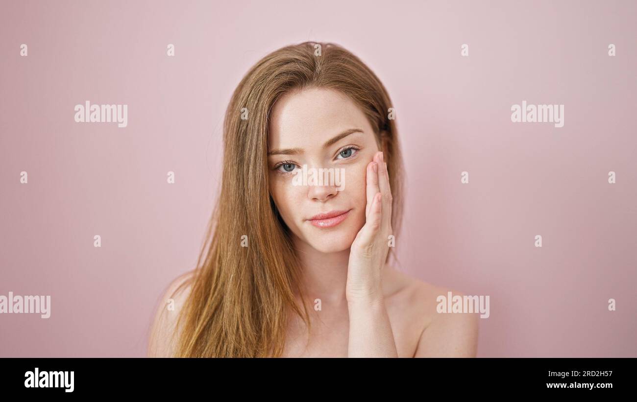 Young blonde woman touching face with hand over isolated pink ...
