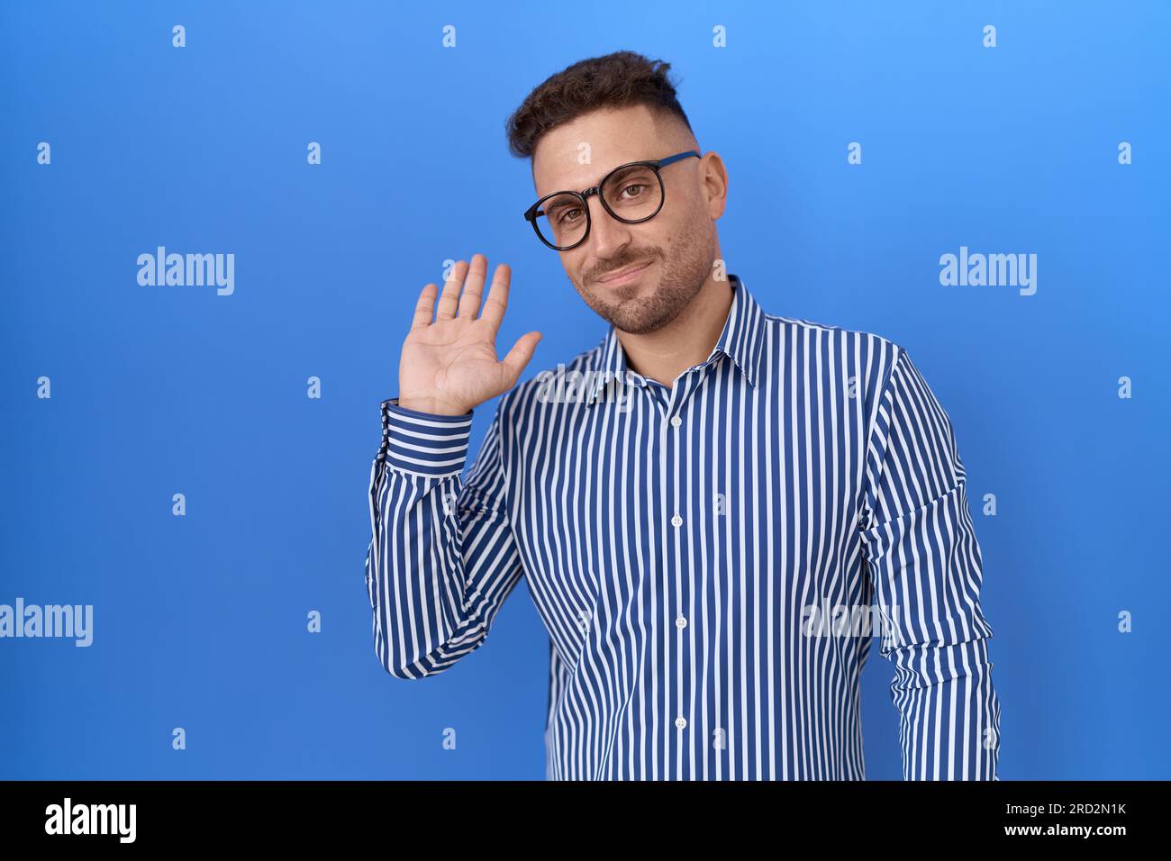 Hispanic man with beard wearing glasses waiving saying hello happy and ...