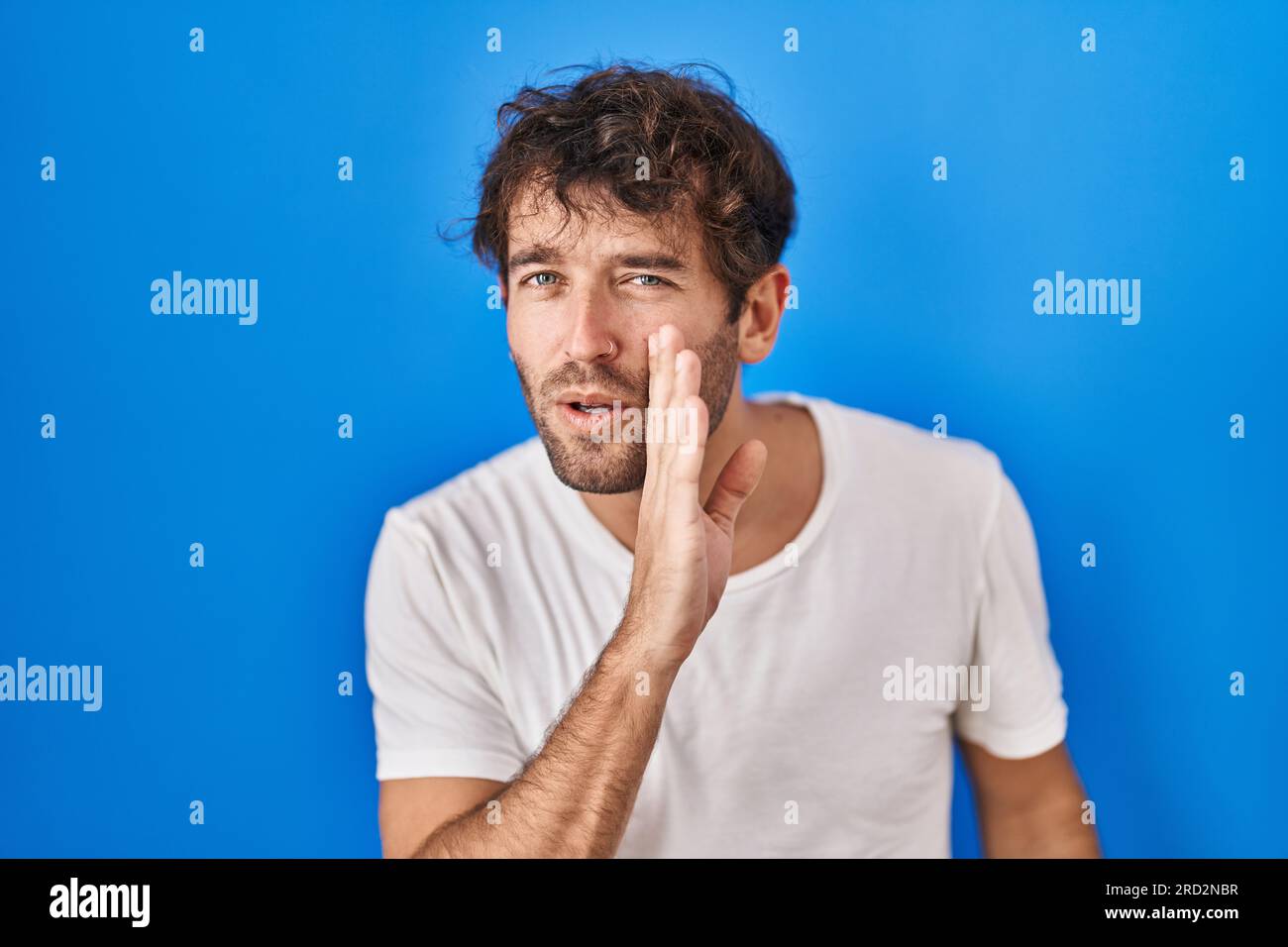 Hispanic young man standing over blue background hand on mouth telling ...