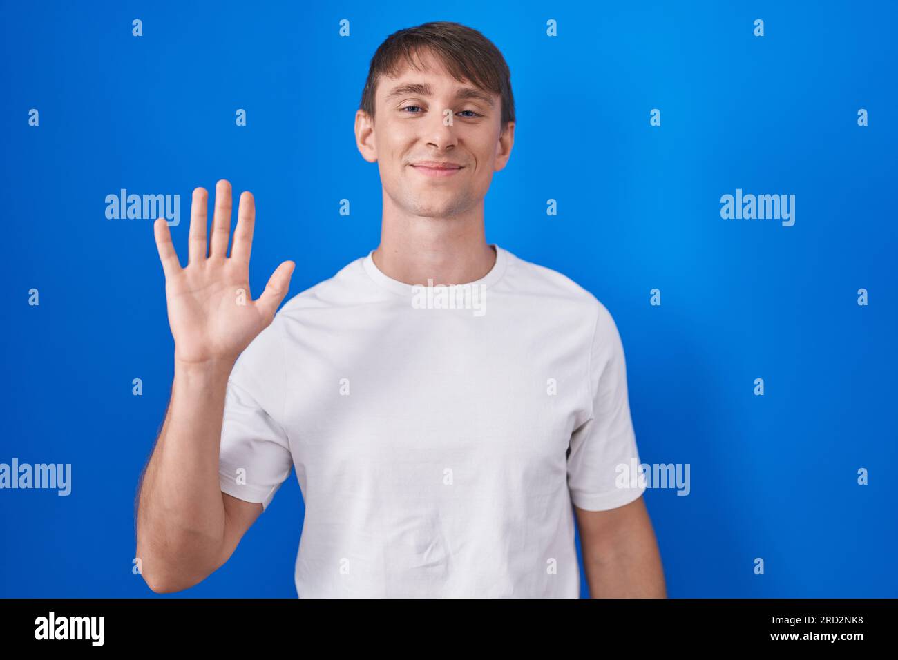 Caucasian blond man standing over blue background waiving saying hello ...