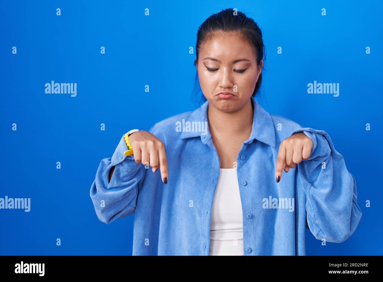 Asian young woman standing over blue background pointing down looking ...