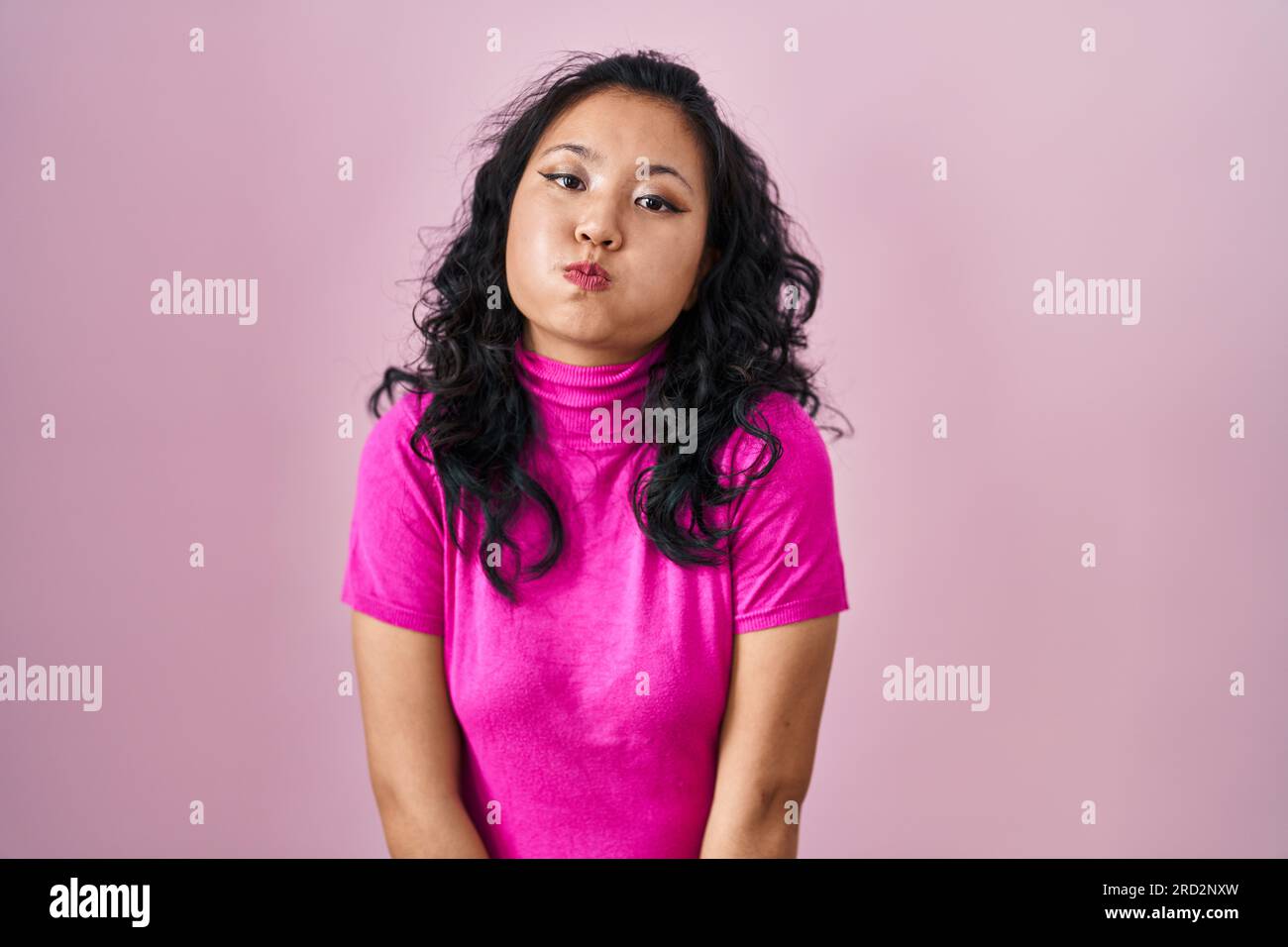 Young asian woman standing over pink background puffing cheeks with ...