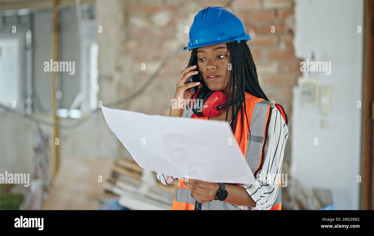 African american woman builder reading house project talking by smartphone at construction site ...