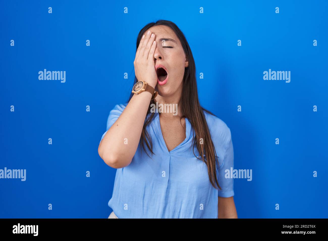 Young brunette woman standing over blue background yawning tired ...