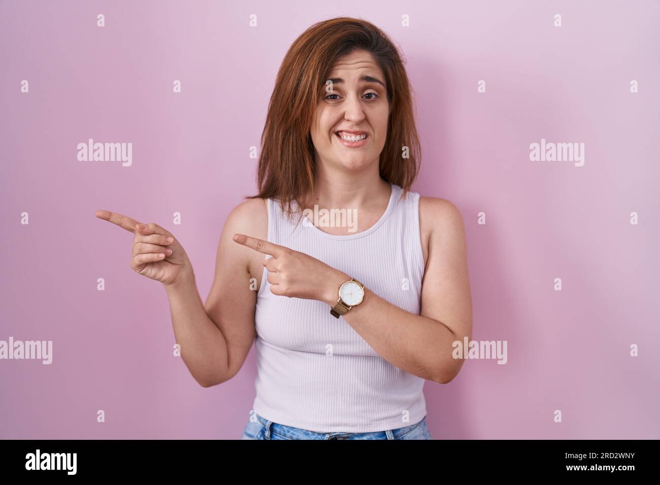 Brunette woman standing over pink background pointing aside worried and ...
