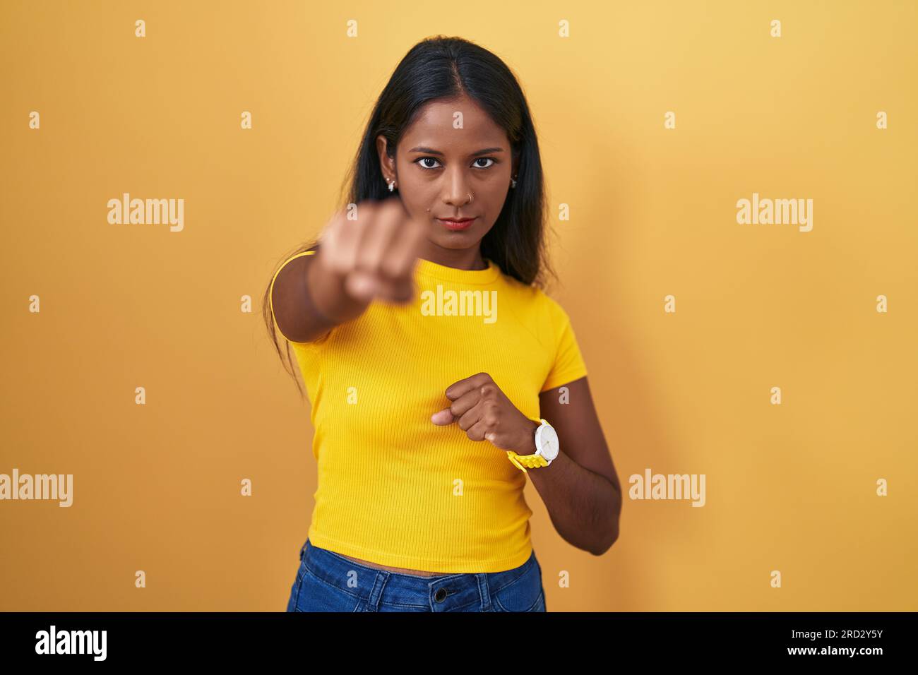 Young indian woman standing over yellow background punching fist to ...