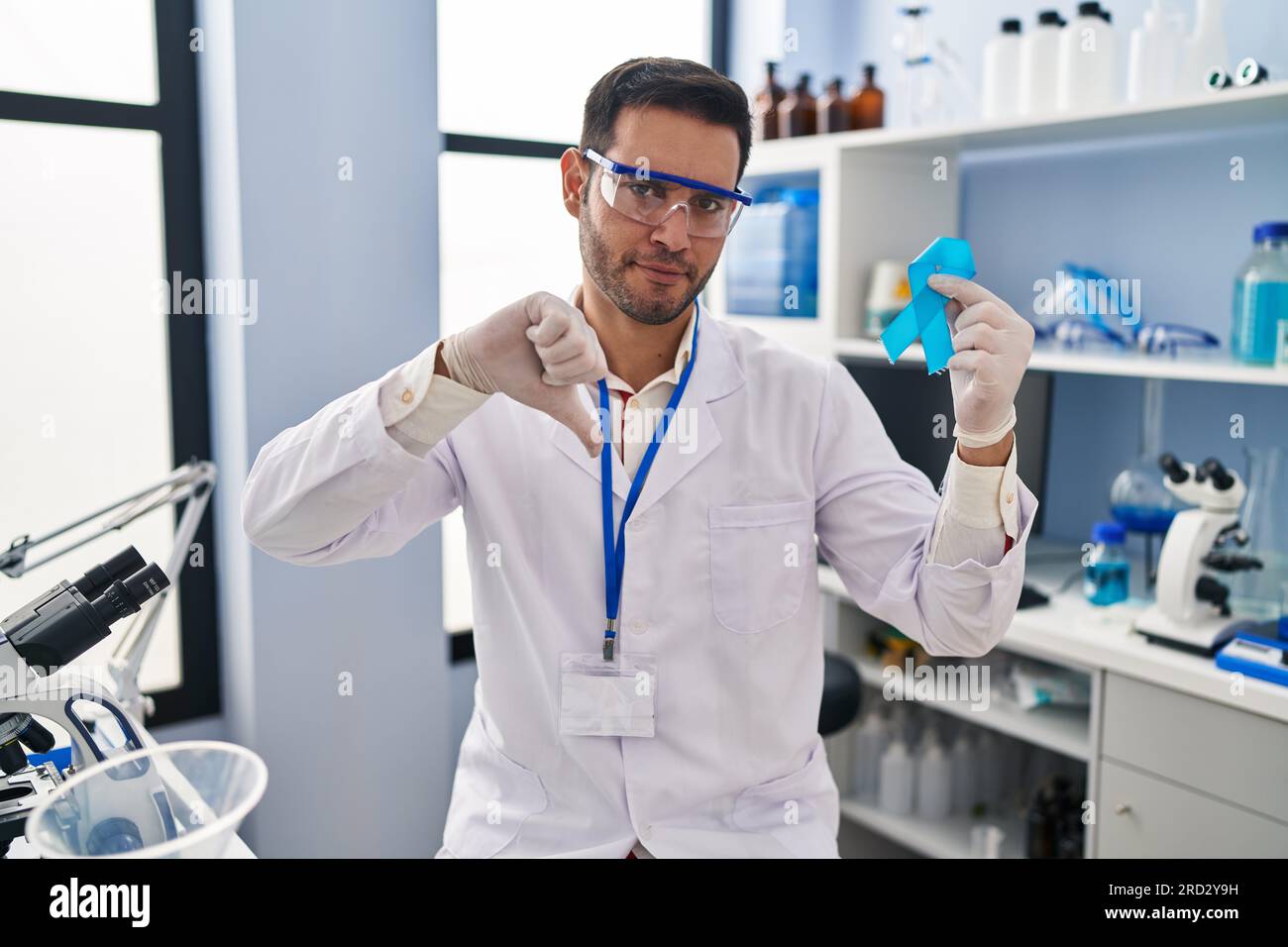 Young hispanic man with beard working at scientist laboratory holding blue ribbon with angry ...