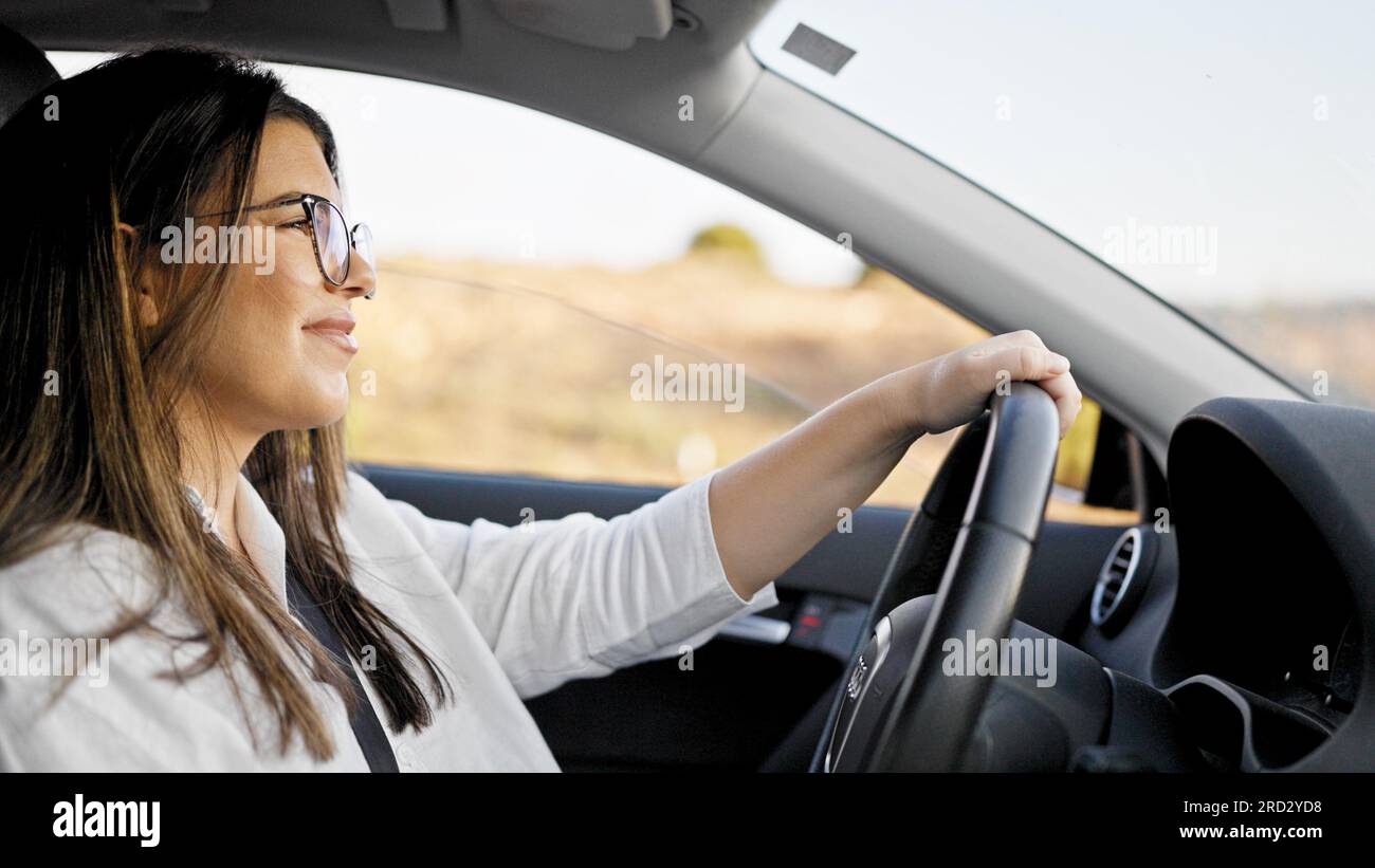 Young beautiful hispanic woman driving a car smiling wearing glasses on ...