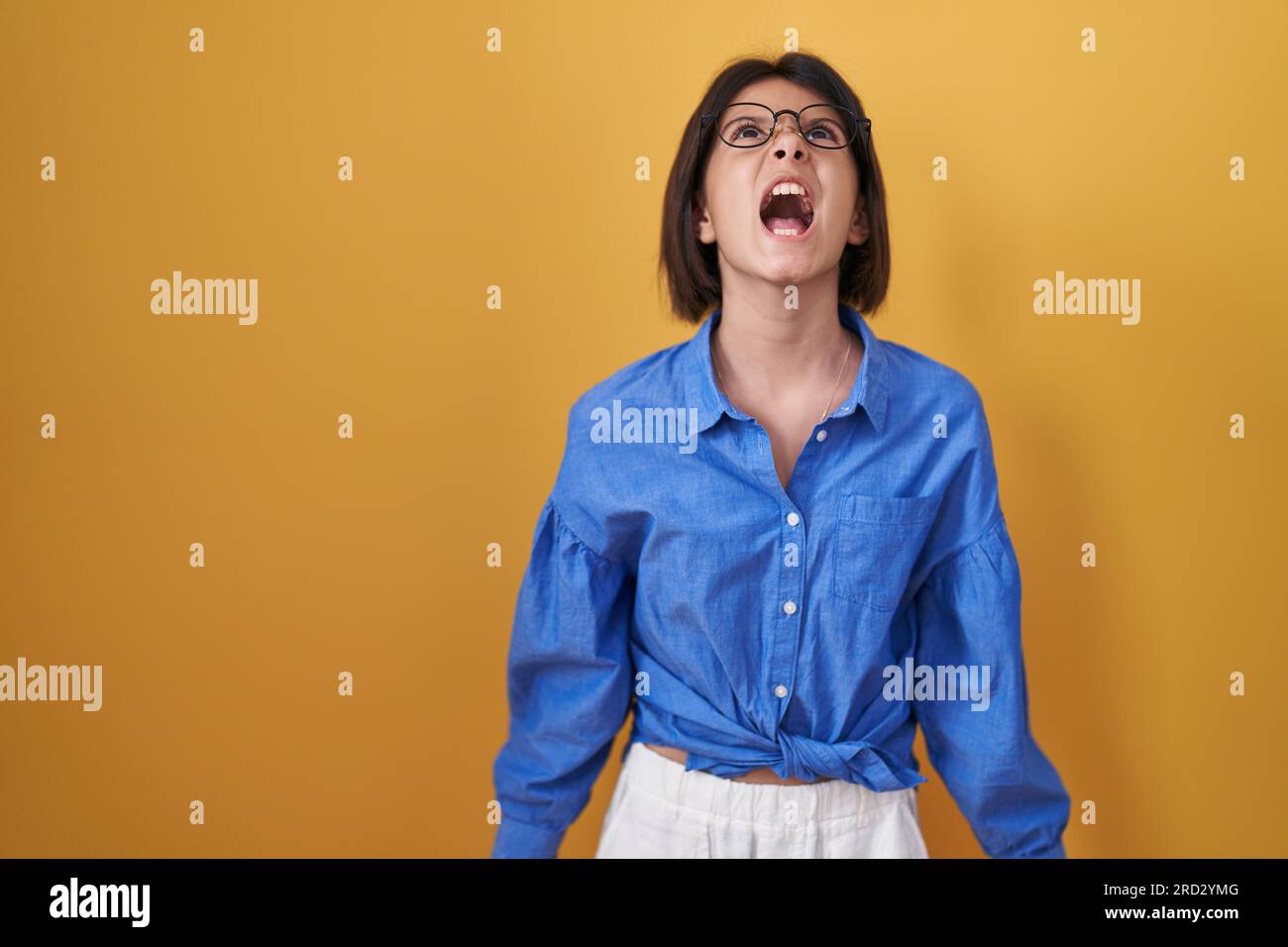 Young girl standing over yellow background angry and mad screaming ...