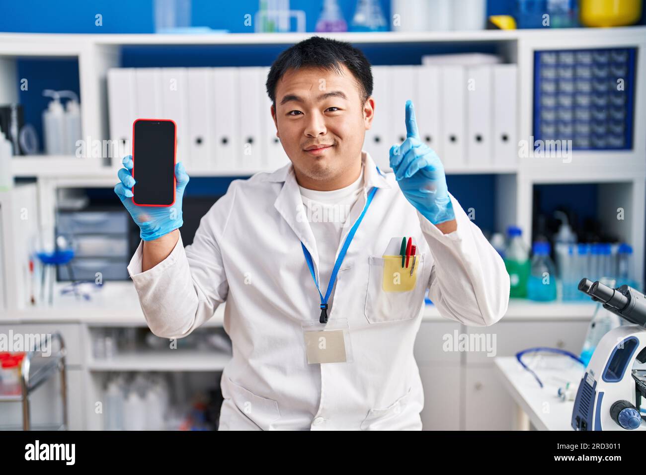 Young chinese man working at scientist laboratory holding smartphone surprised with an idea or ...