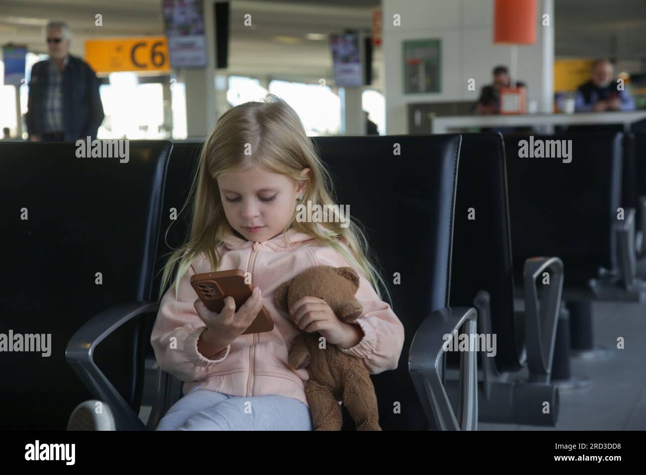 Little girl sitting in an airport departure hall holding teddy bear and ...