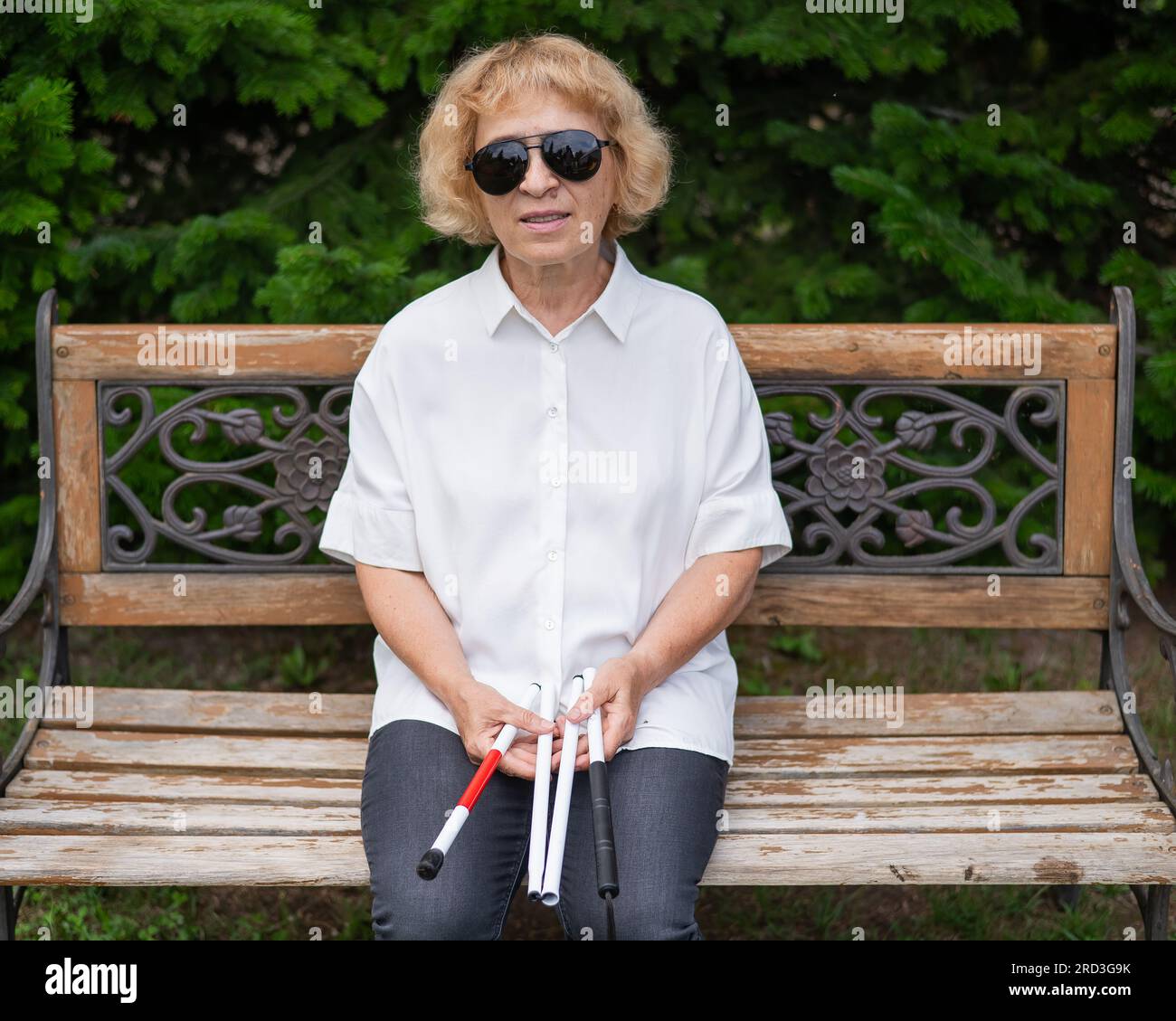 An elderly blind woman sits on a bench in the park with a folded ...