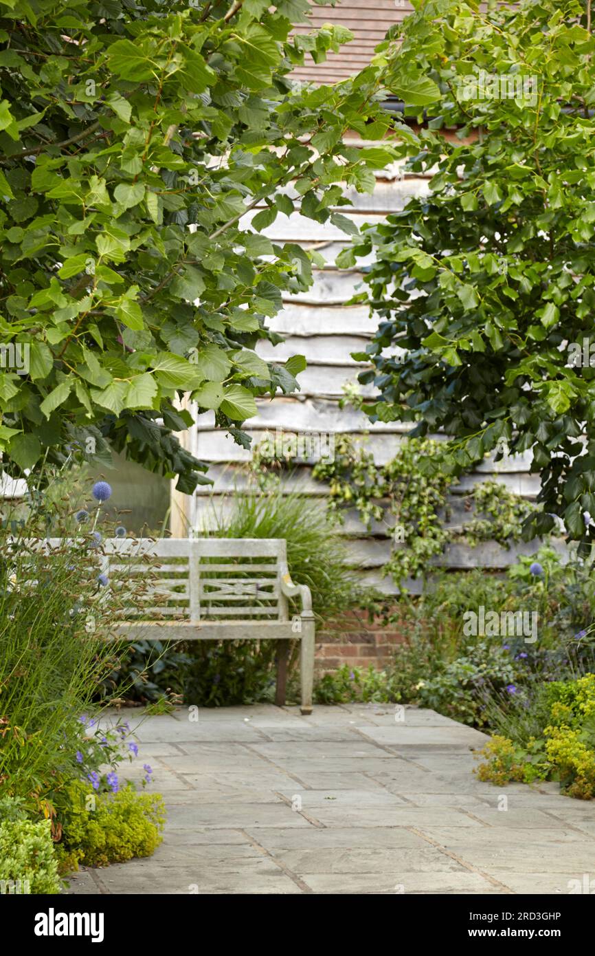 Wood garden bench on paved area in front of stone wall through trees ...