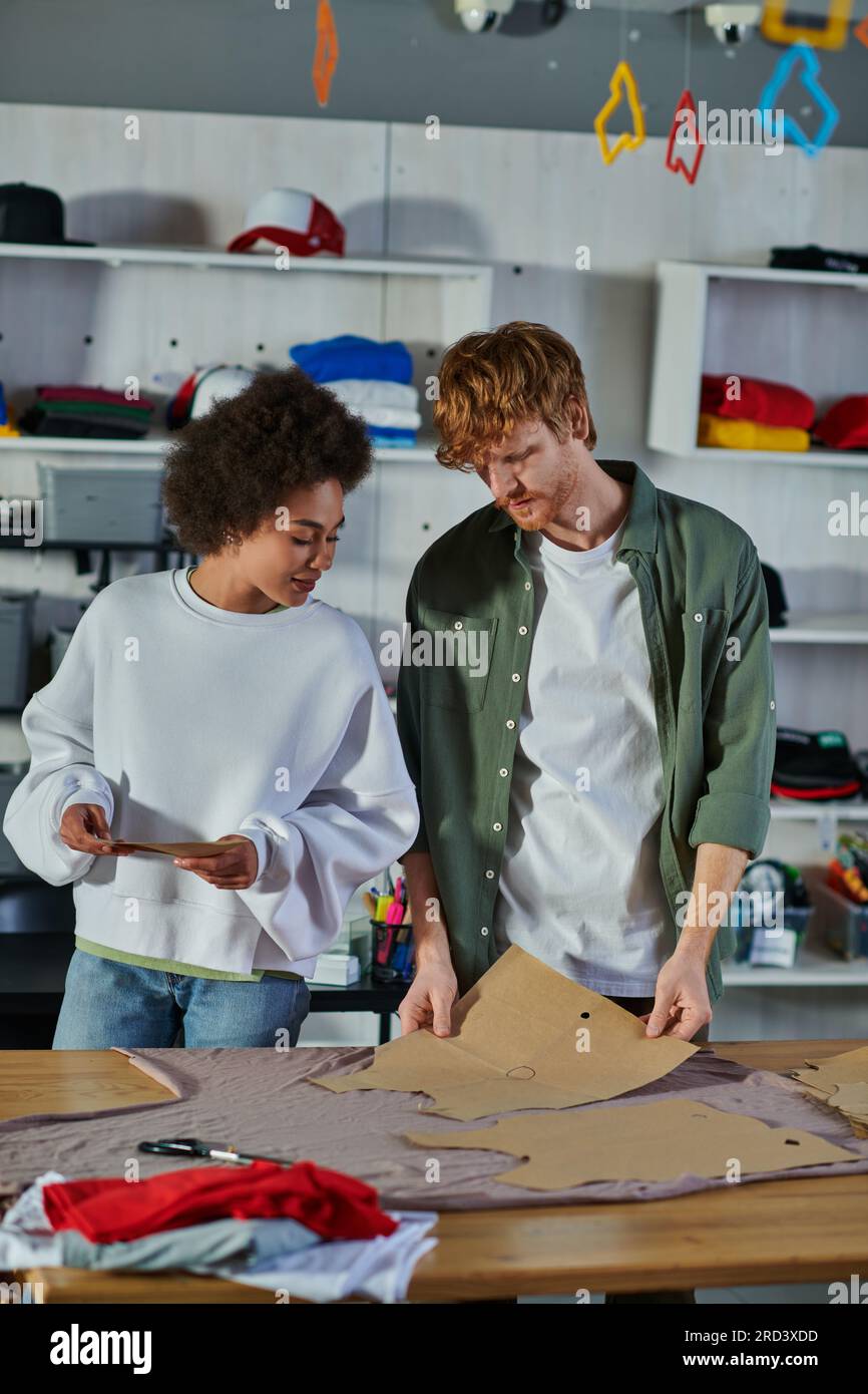 Young redhead craftsman holding sewing pattern near cloth while working ...