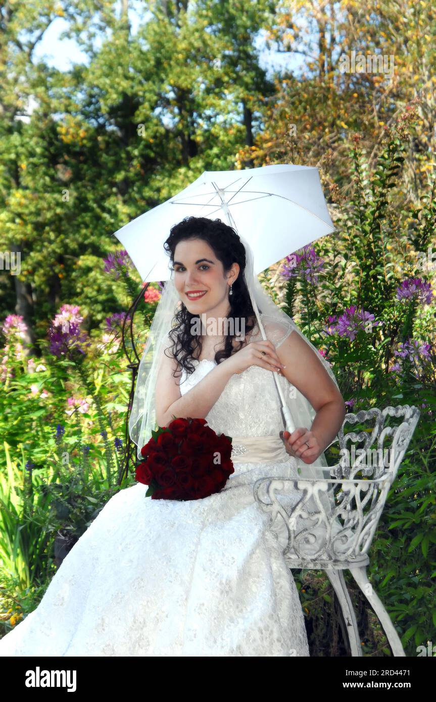 Bride poses, sitting on white, metal chair. She is holding a white ...
