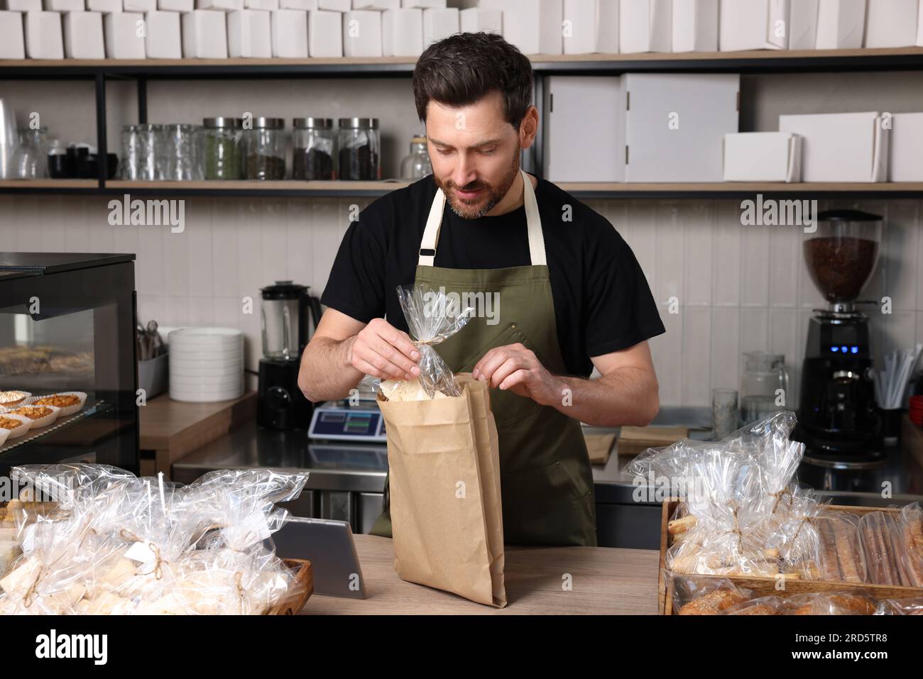 Smiling seller putting pastry into paper bag at cashier desk in bakery ...