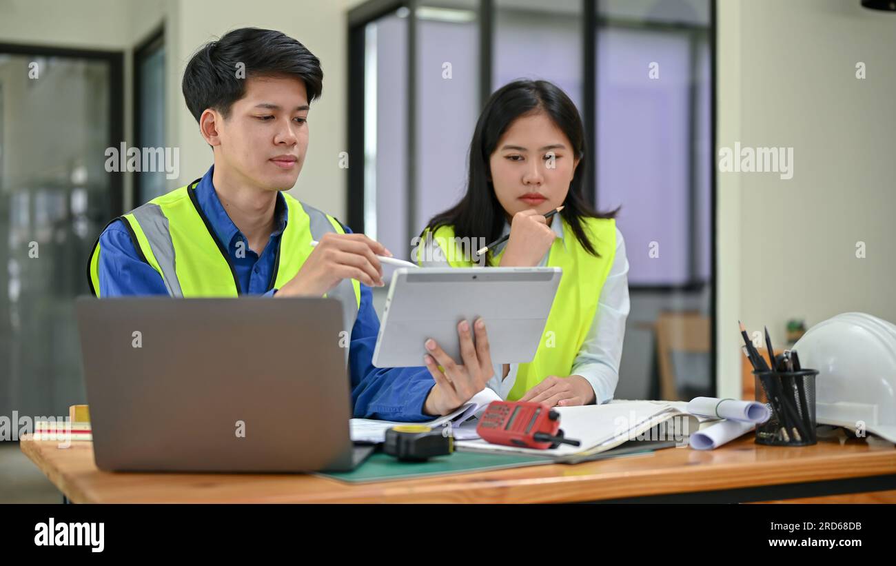 Two professional and serious Asian engineers wearing safety vests are looking at a tablet screen ...
