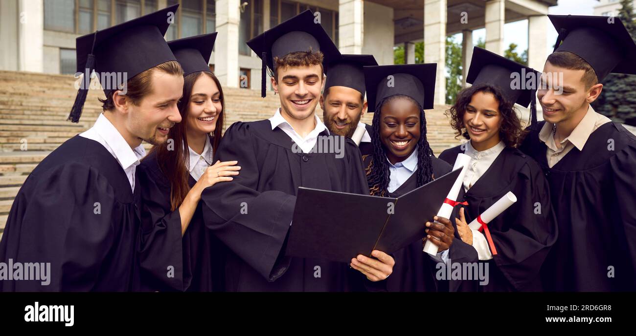 Happy graduate students in black graduation robes looking to diploma of ...