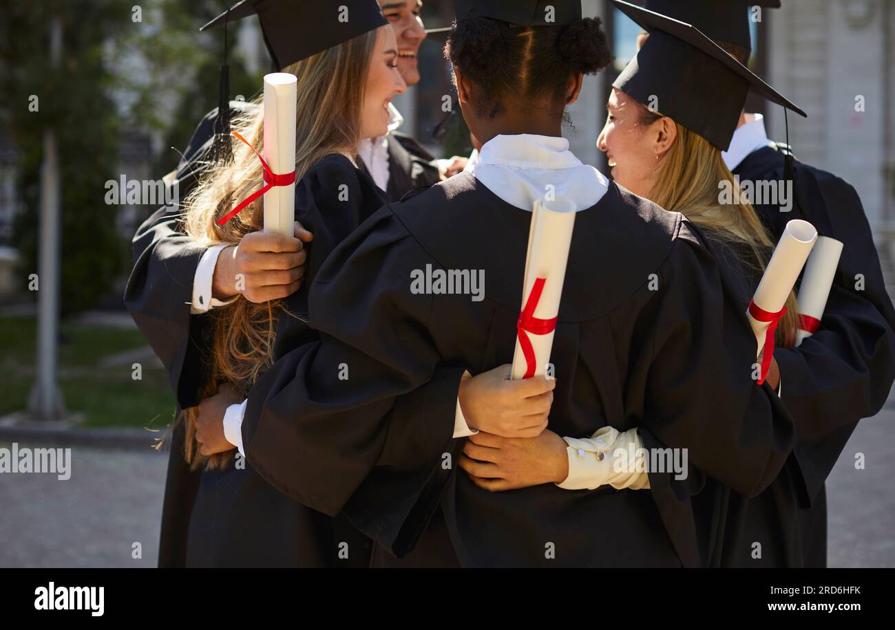 Happy diverse graduates hugging outdoors holding diplomas and ...