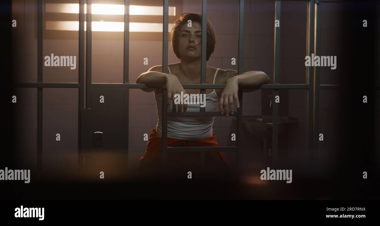 Female prisoner in orange uniform holds hands on bars, stands in jail ...