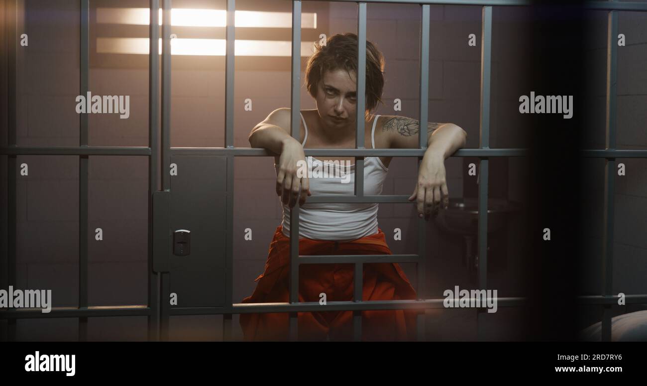 Female prisoner in orange uniform holds hands on bars, stands in jail cell, looks at camera ...