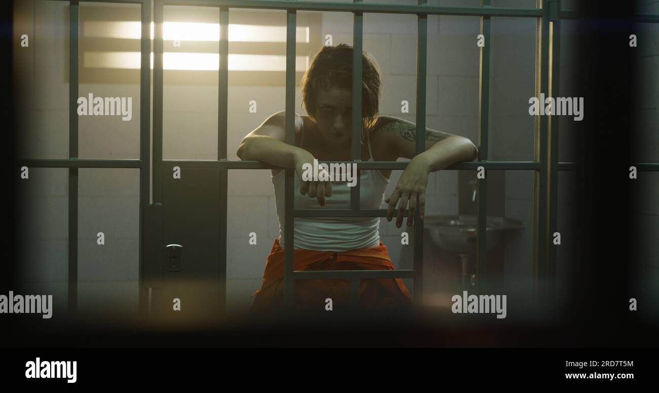 Female prisoner in orange uniform holds hands on bars, stands in jail ...