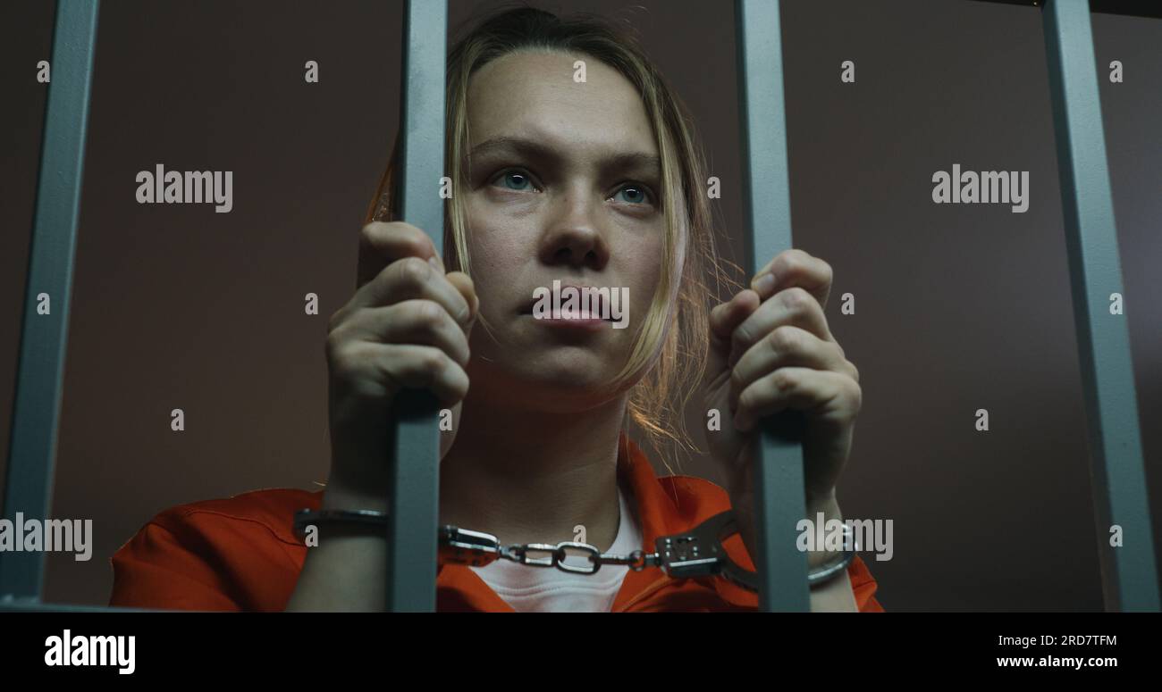Female prisoner in orange uniform holds metal bars, stands in jail cell ...
