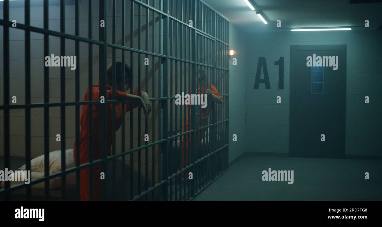 One female prisoner in orange uniform stands behind metal bars, another ...