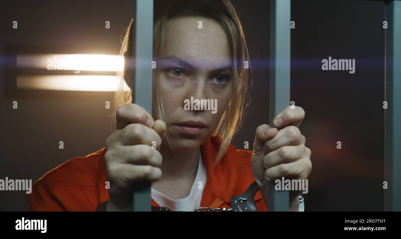 Shocked female prisoner in orange uniform holds metal bars, stands in ...