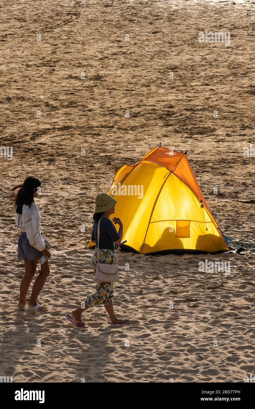 Two holidaymakers walking past a bright yellow beach shelter backlit by ...