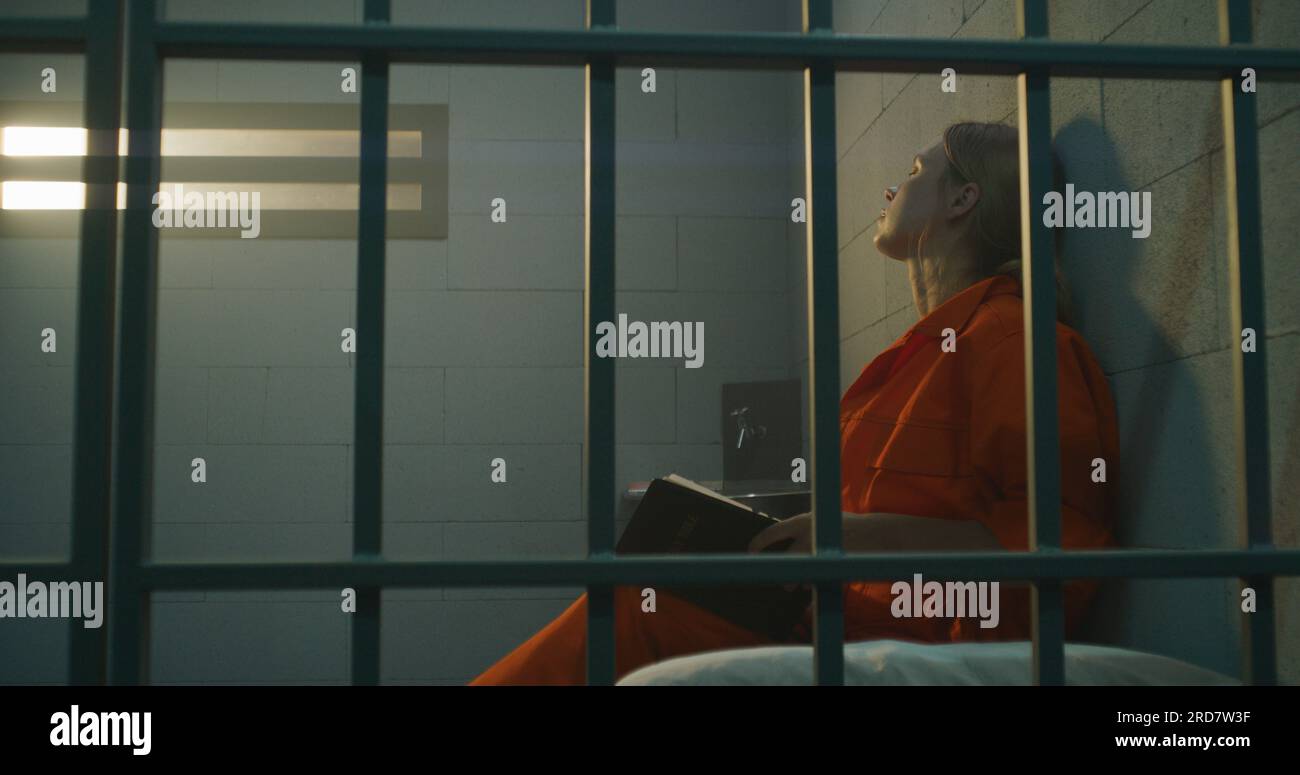 Female prisoner in orange uniform sits on bed behind metal bars, reads ...