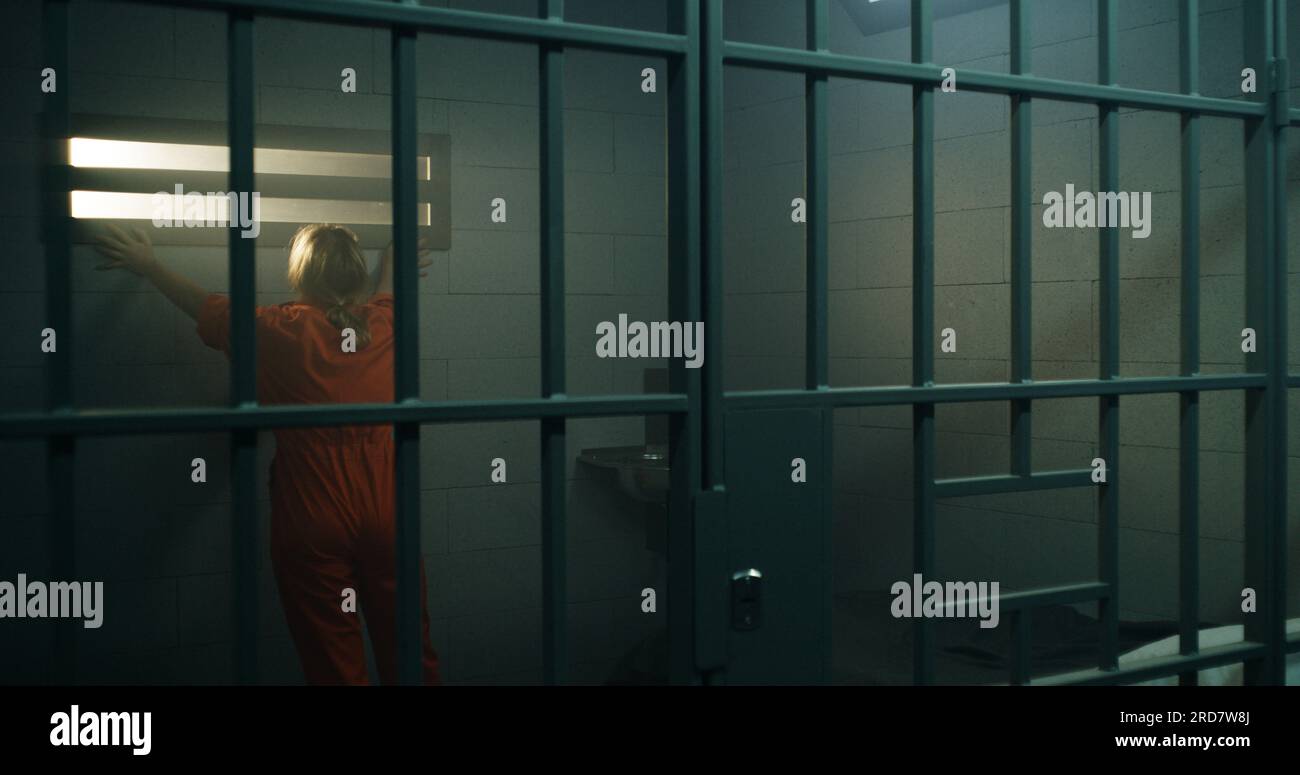 Female prisoner in orange uniform walks in jail cell, looks at barred ...