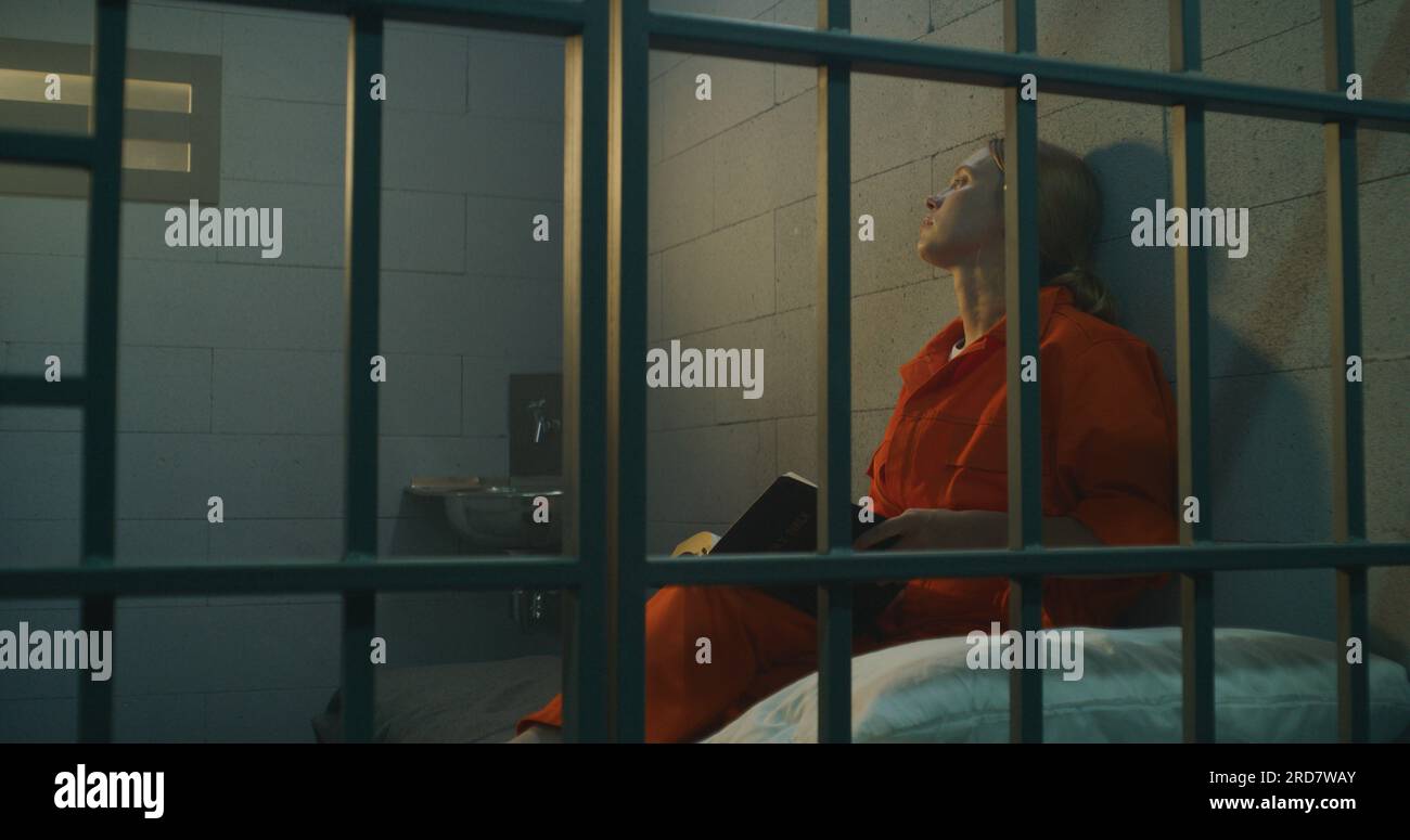 Female prisoner in orange uniform sits on bed behind metal bars, reads ...