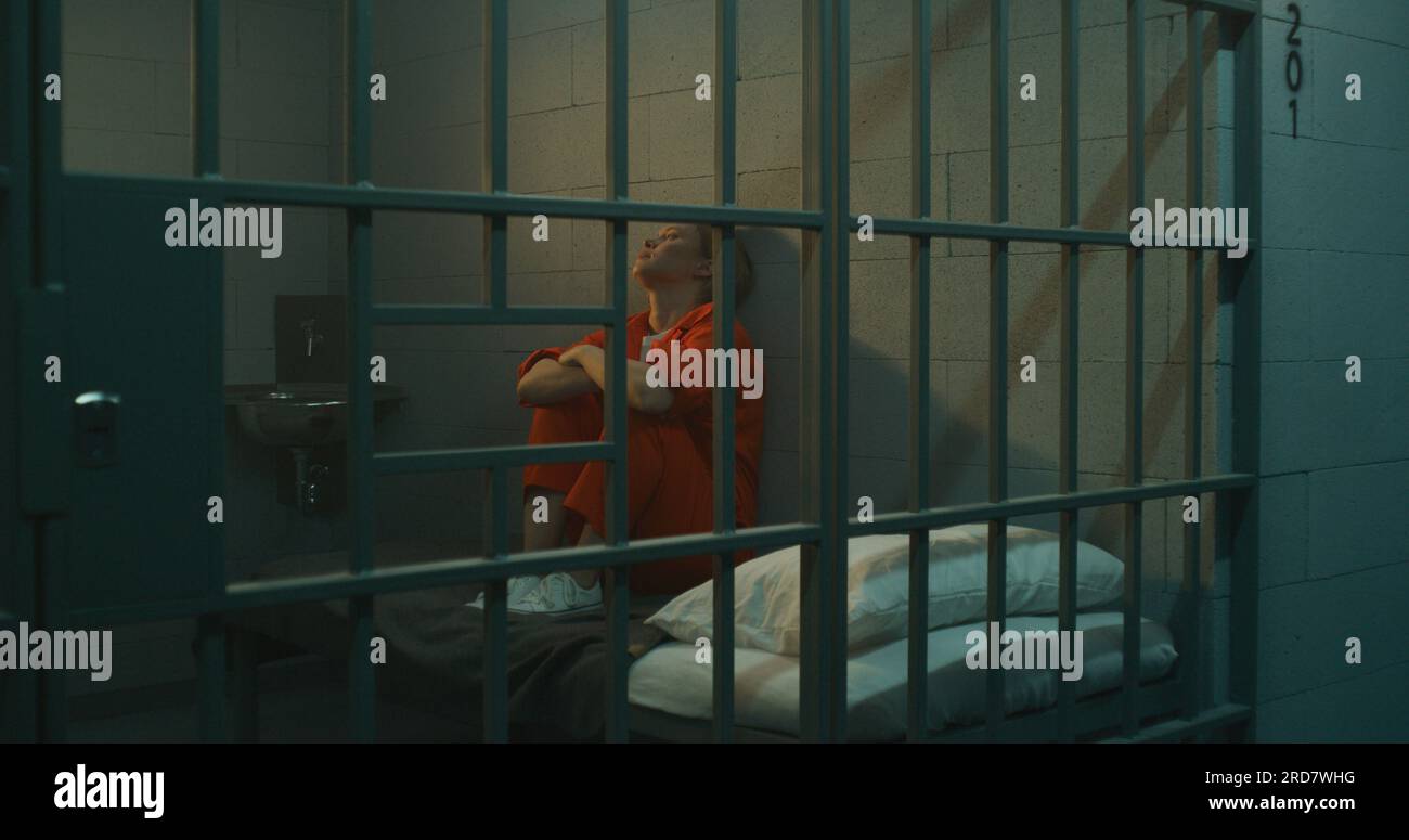 Female prisoner in orange uniform sitting on the bed in jail cell ...