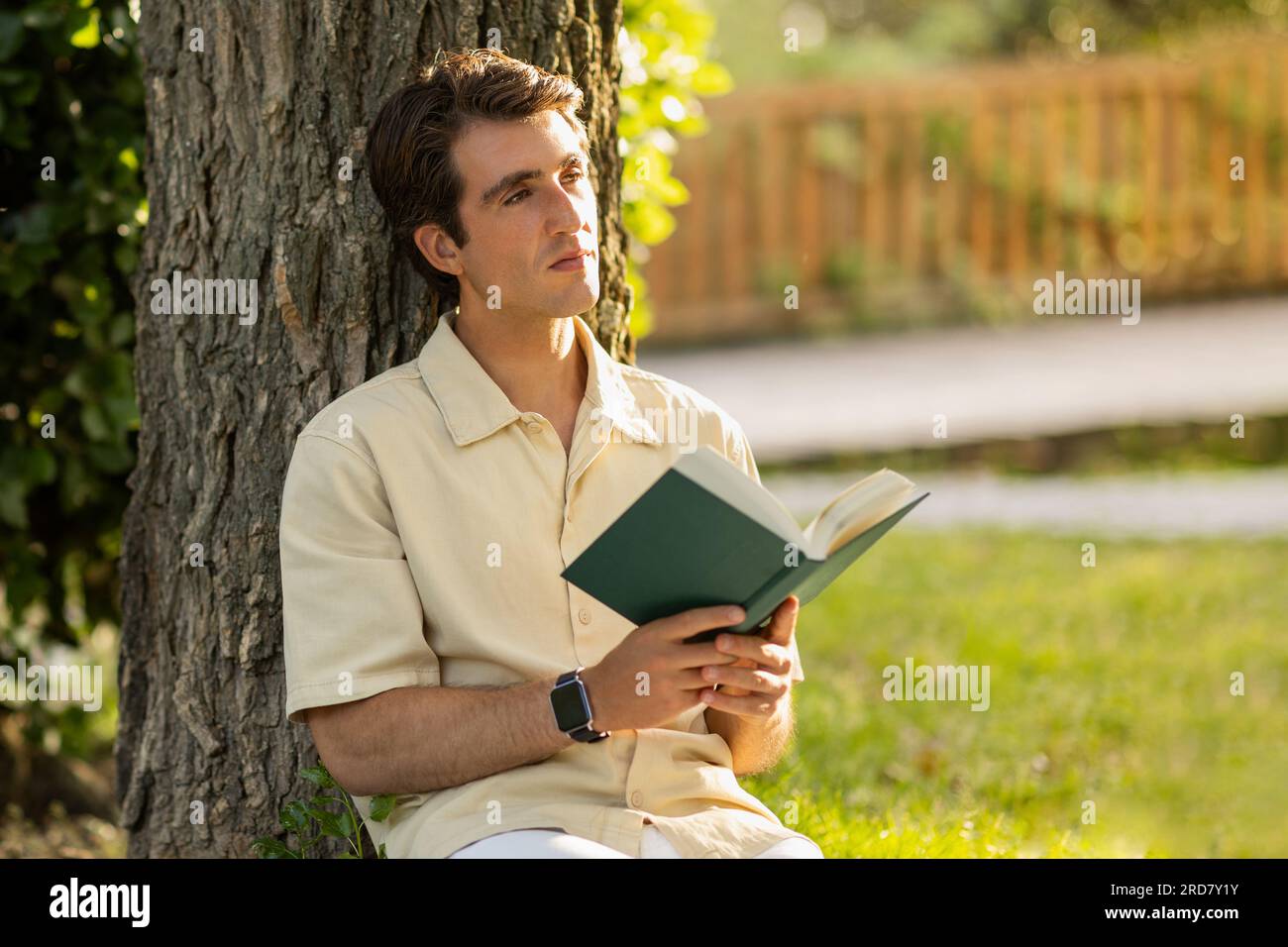 Young guy sitting under tree at garden, reading book Stock Photo - Alamy
