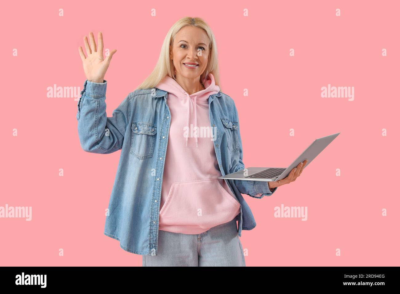 Mature female programmer with laptop waving hand on pink background ...