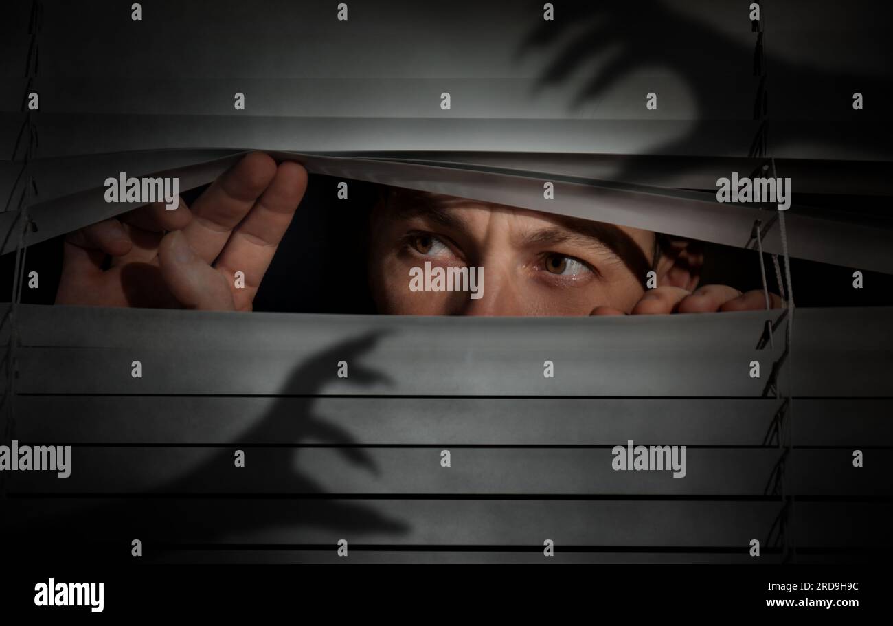 Worried man looking through window blinds into darkness. Shadow of ...