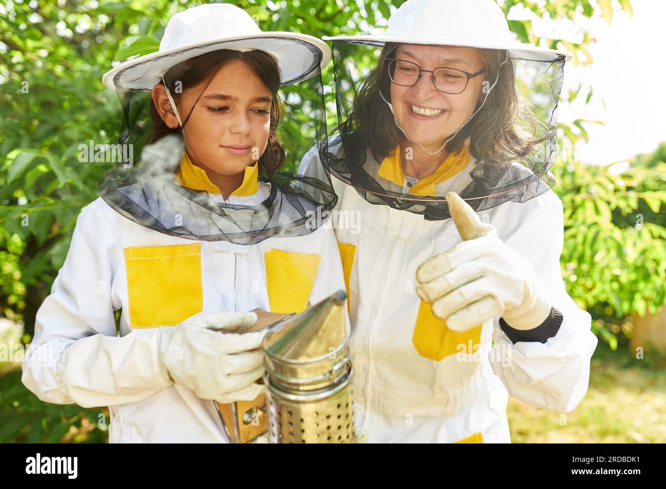 Happy senior female apiarist with girl holding smoker in apiary garden ...