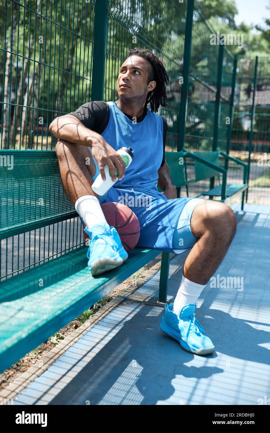 Young man with dreads in blue uniform sitting on bench after playing ...