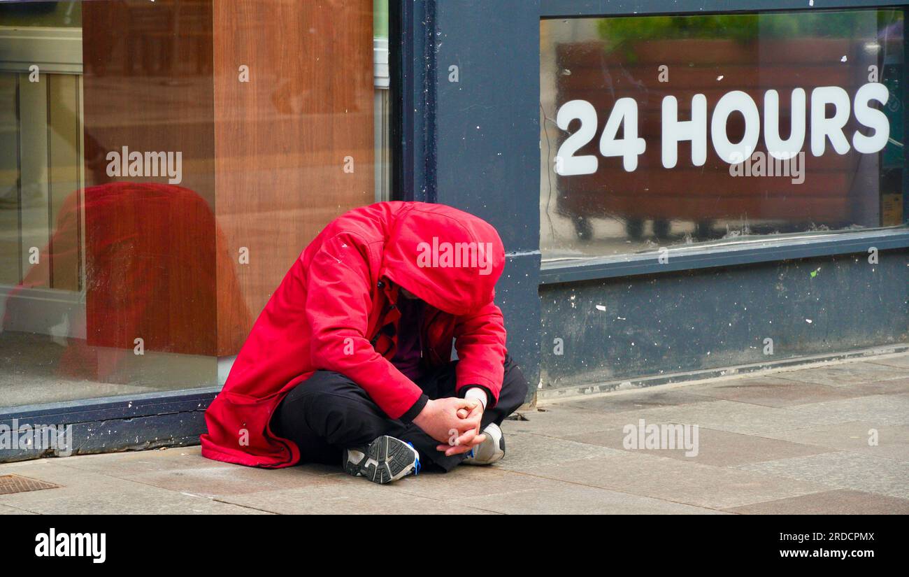 A young person sat on a street pavement curled up in a red hooded ...